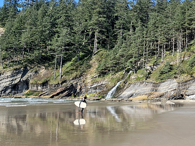Reflections of determination – a lone surfer carries their board across mirror-like wet sand, chasing waves beneath majestic coastal cliffs.