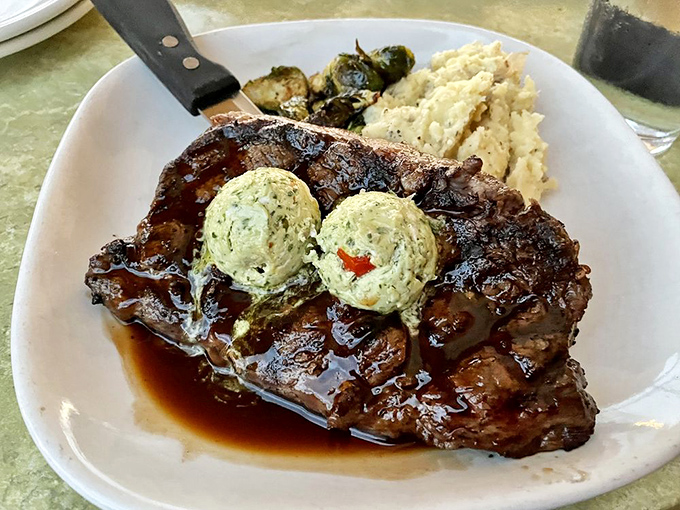 A ribeye steak glistening with savory perfection, topped with herb butter melting into a pool of deliciousness. Meat poetry on a plate.
