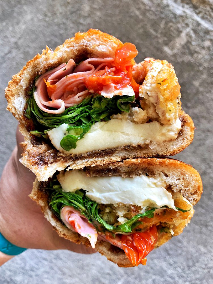 A sandwich cross-section revealing layers of delicious complexity. Roasted red peppers, fresh greens, thinly sliced meats, and melty cheese create a masterpiece of flavor architecture.