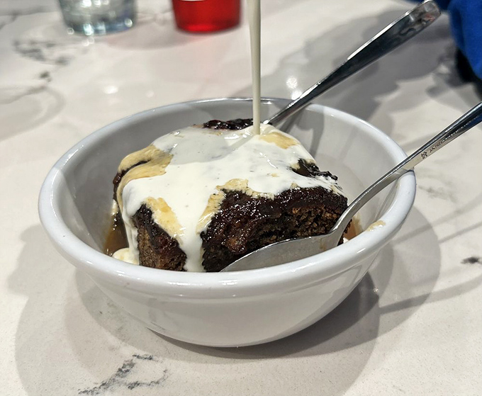 Sticky toffee pudding with cream being poured over like a warm British hug in dessert form—resistance is absolutely futile.