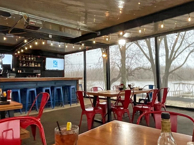 String lights dance overhead as red metal chairs pop against the blue bar, all while floor-to-ceiling windows frame the lake view like nature's television.