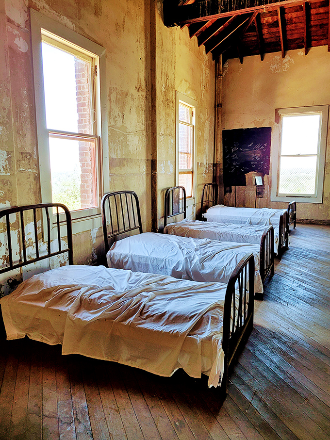 The dormitory's spartan iron beds lined up with military precision tell a stark tale of institutional life, their crisp white sheets a ghostly contrast to weathered walls.