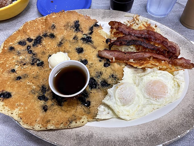 The legendary blueberry pancake stretches to the edge of the plate, dotted with berries like a delicious constellation, accompanied by perfectly crisp bacon.