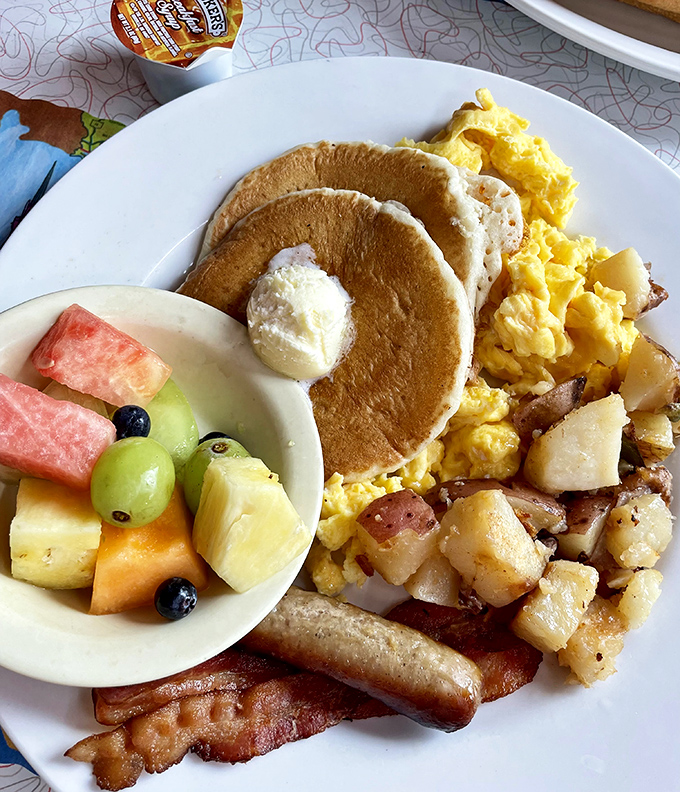 Breakfast nirvana on a plate: golden pancakes, scrambled eggs, and crispy bacon sharing space with fresh fruit that pretends this is a balanced meal.