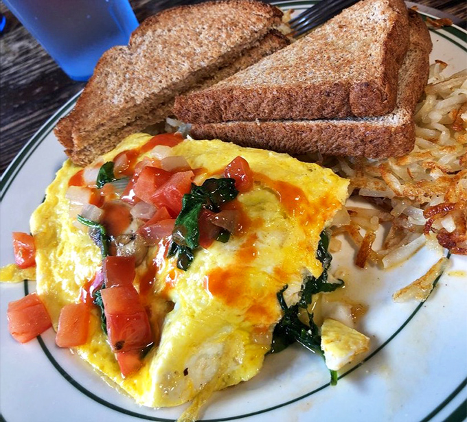 Breakfast artistry at its finest&mdash;a vibrant omelet bursting with fresh spinach and tomatoes alongside perfectly crisped hash browns and hearty toast.