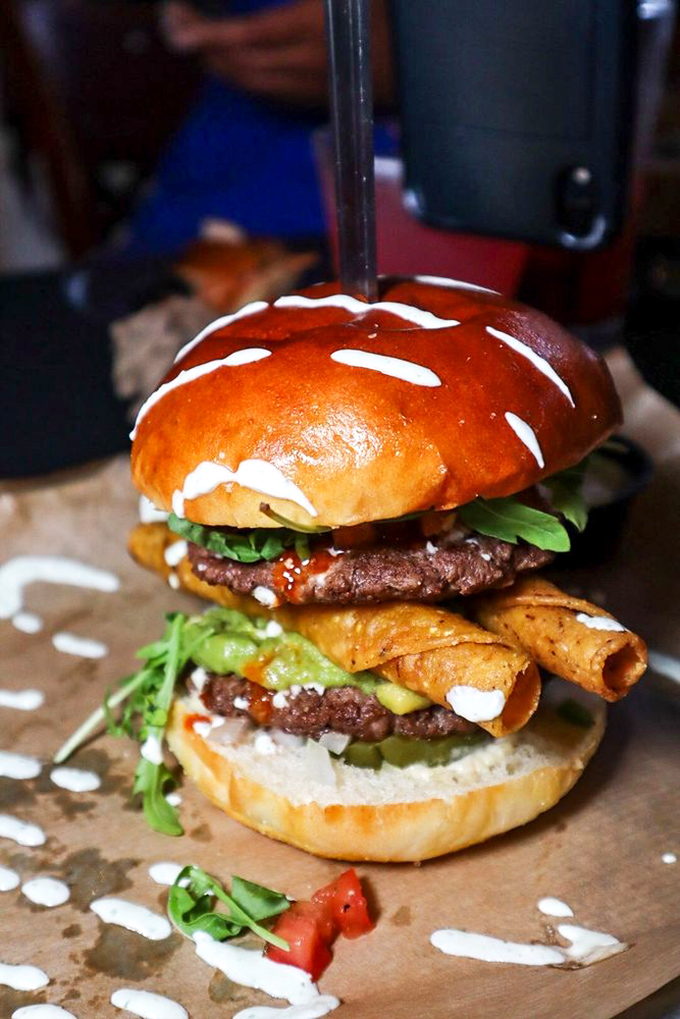 Architectural masterpiece or burger? Why not both! This towering creation with taquitos and dual patties defies both gravity and conventional sandwich logic.