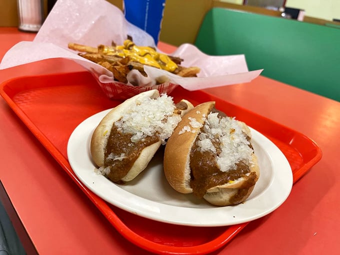 Two perfect chili dogs nestled on a simple white plate. The chopped onions add that essential crunch while the sauce whispers, "This is why you came."
