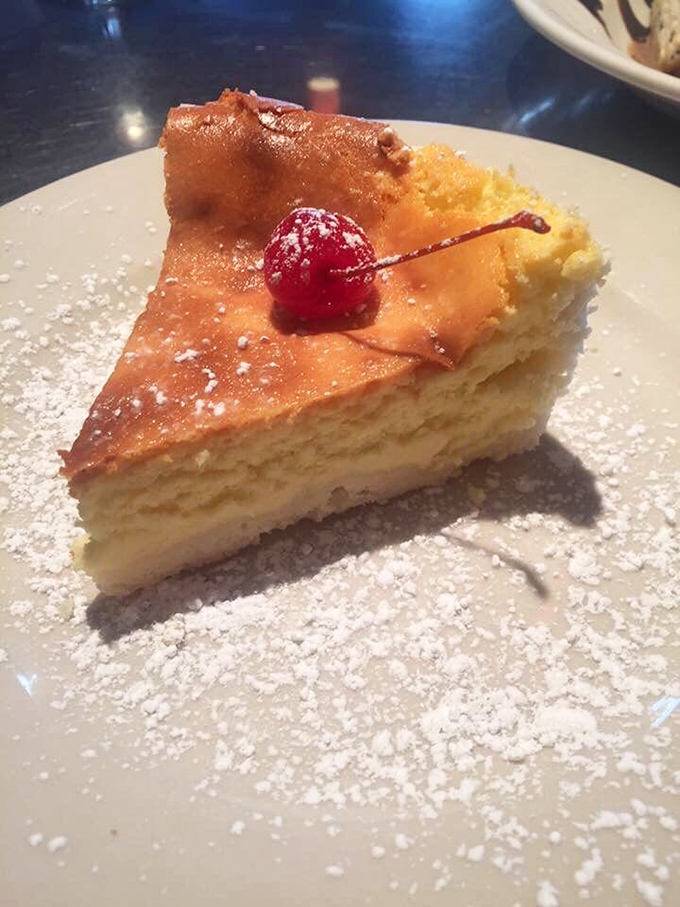 The Italian cheesecake wears its golden top like a crown, dusted with powdered sugar snow and crowned with a cherry jewel.