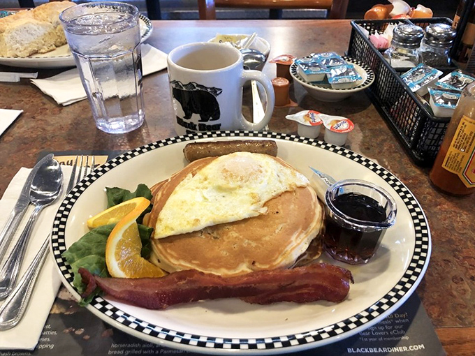 The perfect breakfast trifecta: a golden pancake, crispy bacon, and a sunny-side egg, all served on the diner's signature checkered plates that scream "comfort food ahead!"