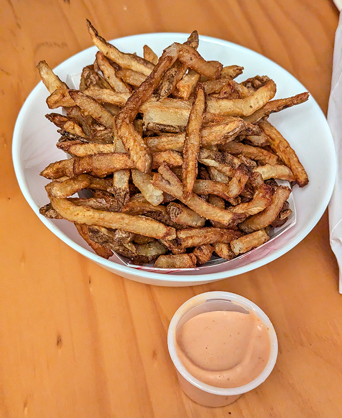 A pristine white bowl showcases these hand-cut fries like the treasures they are. That little cup of fry sauce? The perfect dance partner for potato perfection.