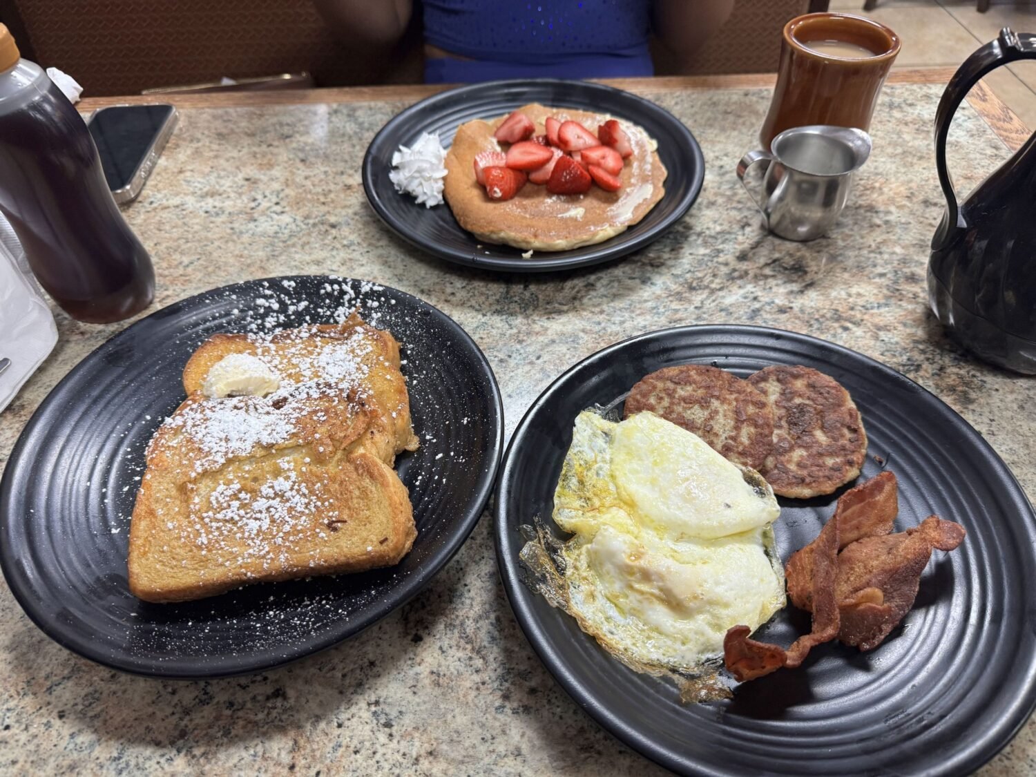 Breakfast perfection on black plates - French toast dusted with powdered sugar, eggs with perfectly crisp edges, and a pancake that's practically begging for maple syrup.