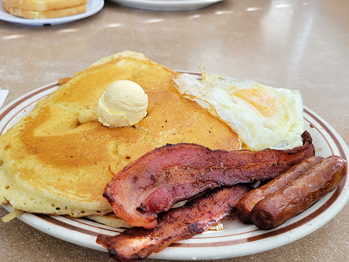 The breakfast trinity—perfect pancake, crispy bacon, and sunny-side-up egg—arranged on a plate like old friends at a reunion. Pure morning magic.