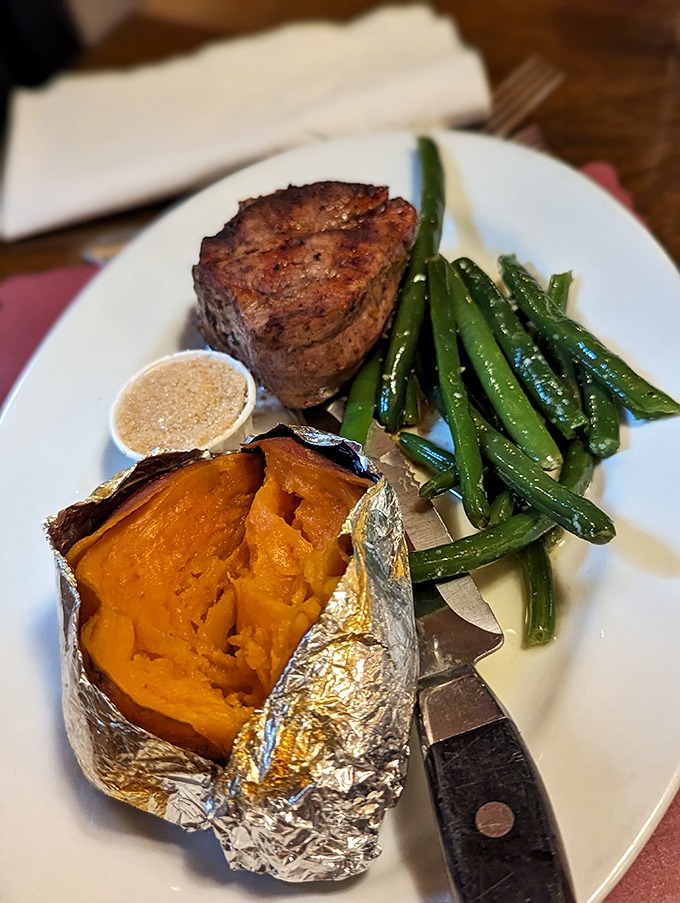 A perfectly seared filet mignon stands guard beside a sweet potato bursting from its foil jacket—proof that simple ingredients need only proper respect.