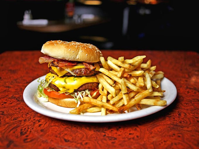 This towering burger masterpiece with its cascade of melted cheese and crispy bacon sits beside golden fries like royalty holding court on a white plate.