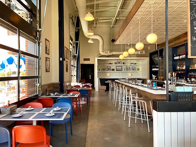 The airy interior combines industrial elements with coastal comfort. High ceilings, pendant lighting, and those signature blue and red chairs create a relaxed vibe.