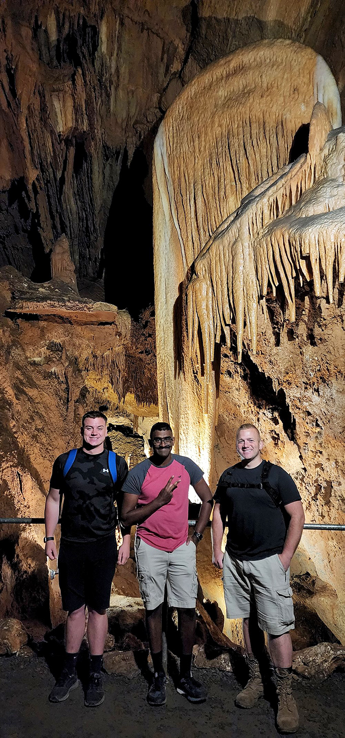 Three friends pose beneath a towering flowstone curtain, providing scale that highlights the cavern's impressive dimensions.