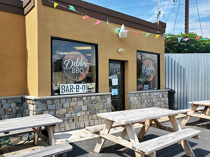 The unassuming tan exterior of Dibbs BBQ, where colorful pennant flags and wooden picnic tables hint at the smoky treasures waiting inside.
