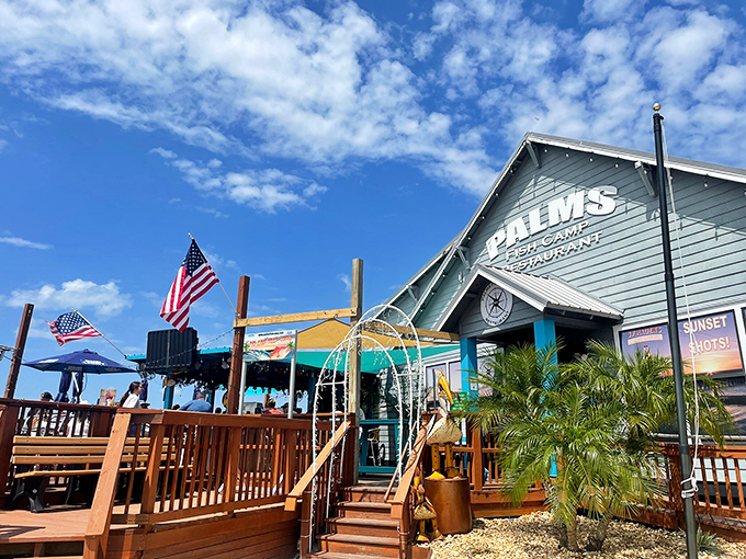 The blue-hued exterior of Palms Fish Camp Restaurant stands like a beacon for seafood lovers, with American flags fluttering above the wooden deck where waterfront dreams come true.