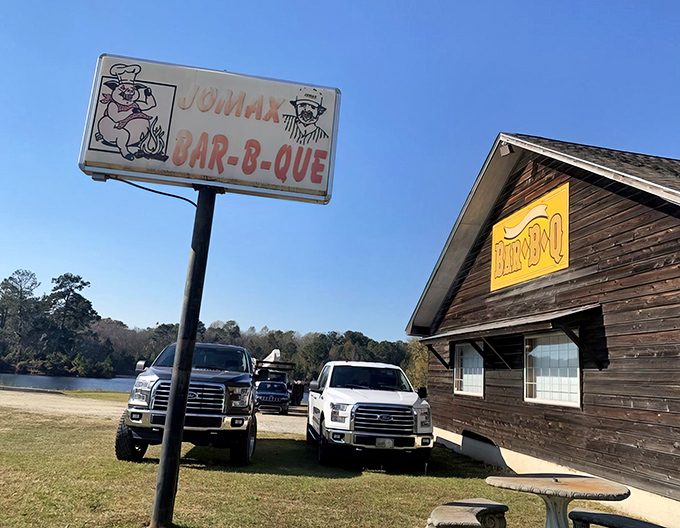 The weathered wooden exterior of Jomax BBQ stands like a barbecue time capsule against Georgia's blue sky, promising smoky treasures within.