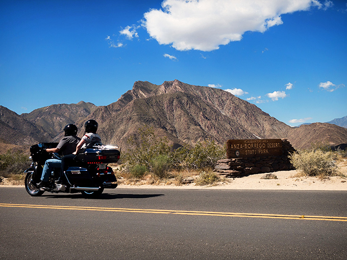 The open road beckons as motorcyclists cruise past rugged mountain terrain, where adventure awaits around every bend at Anza-Borrego's magnificent entrance.