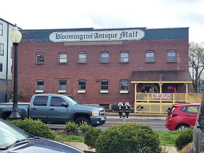 The brick facade of Bloomington Antique Mall stands like a time capsule with its cheerful yellow porch proudly announcing "WE ARE OPEN!" &ndash; an invitation to adventure.