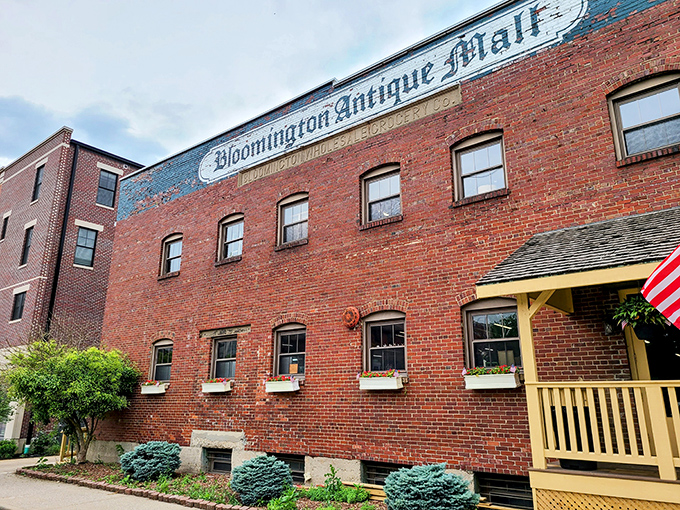 The brick facade of Bloomington Antique Mall stands like a time capsule with its cheerful yellow porch proudly announcing "WE ARE OPEN!" &ndash; an invitation to adventure.