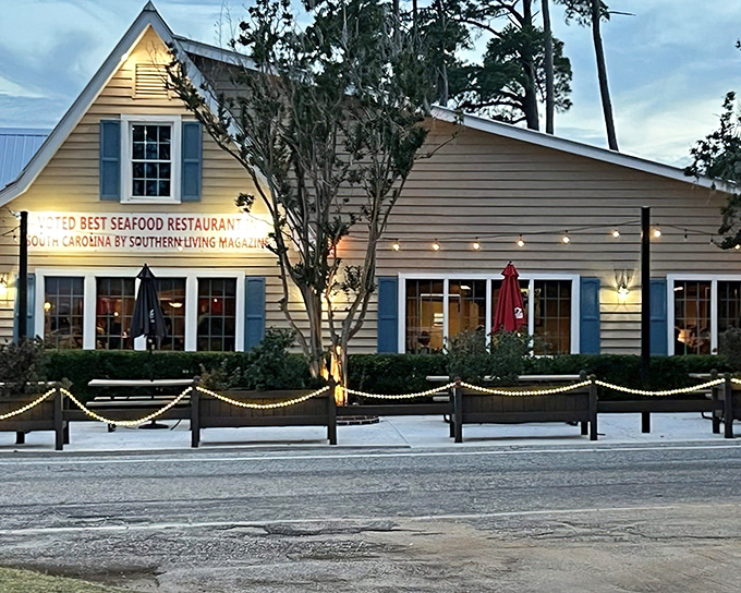 The charming yellow exterior of Lee's Inlet Kitchen glows at dusk, a beacon for seafood lovers with its Southern Living endorsement proudly displayed.