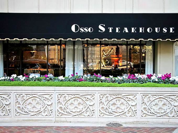Elegance meets urban charm at Osso Steakhouse's inviting exterior, where flowering plants and a classic awning hint at the culinary delights waiting inside.