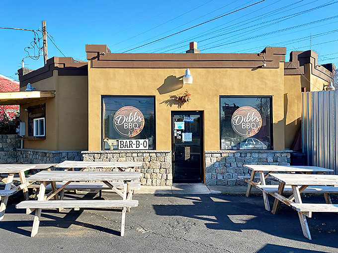 The unassuming tan exterior of Dibbs BBQ, where colorful pennant flags and wooden picnic tables hint at the smoky treasures waiting inside.