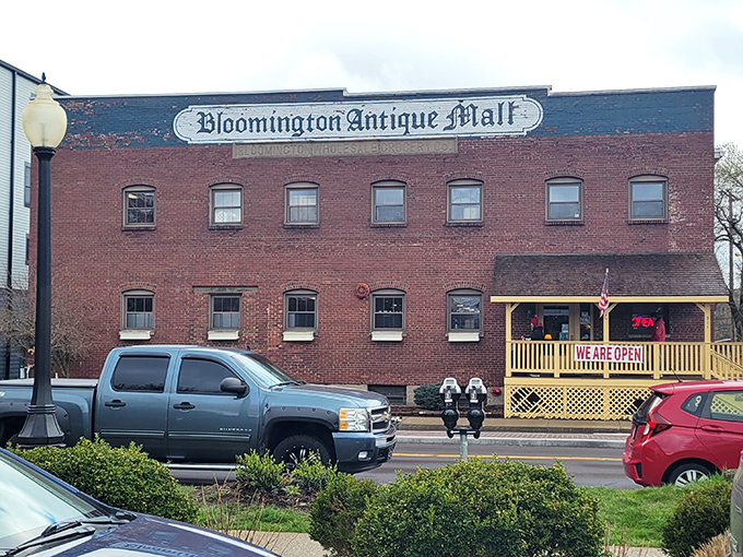 The brick facade of Bloomington Antique Mall stands like a time capsule with its cheerful yellow porch proudly announcing "WE ARE OPEN!" &ndash; an invitation to adventure.