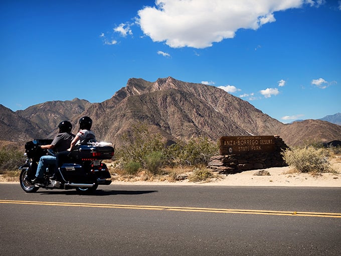The open road beckons as motorcyclists cruise past rugged mountain terrain, where adventure awaits around every bend at Anza-Borrego's magnificent entrance.