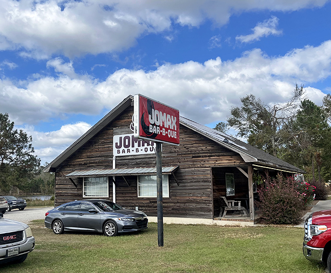 The weathered wooden exterior of Jomax BBQ stands like a barbecue time capsule against Georgia's blue sky, promising smoky treasures within.