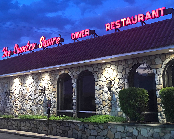 The neon glow of Country Squire Diner's sign against twilight skies promises comfort food salvation after a long day. Stone walls and warm lighting beckon hungry travelers inside.