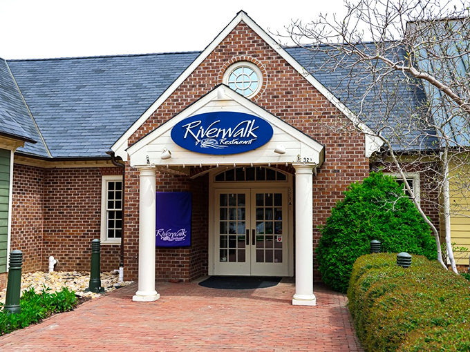 The welcoming brick fa&ccedil;ade of Riverwalk Restaurant stands like a culinary lighthouse, beckoning hungry travelers with its classic colonial charm and blue signage.