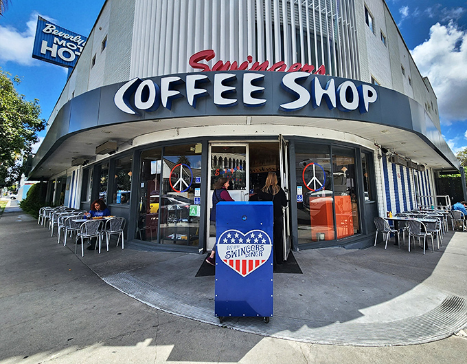 The iconic corner entrance of Swingers, where the neon sign and peace symbols promise both nostalgia and pancakes worth crossing Beverly Boulevard for.