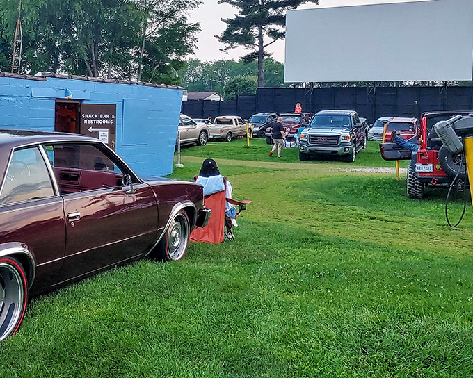 Cars line up at dusk, their headlights dimmed in anticipation. The blue concession building stands ready to serve moviegoers seeking that perfect summer night.