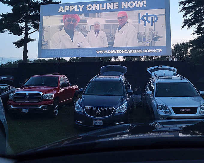 Cars line up at dusk, their headlights dimmed in anticipation. The blue concession building stands ready to serve moviegoers seeking that perfect summer night.