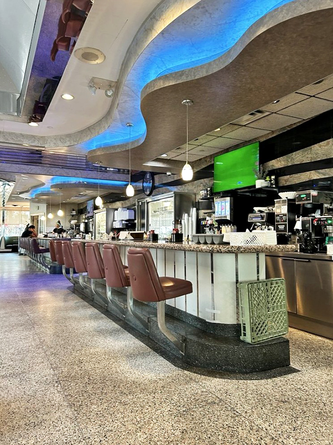 The counter beckons with its row of burgundy stools, where solo diners can perch and watch the kitchen ballet unfold beneath soft pendant lighting.