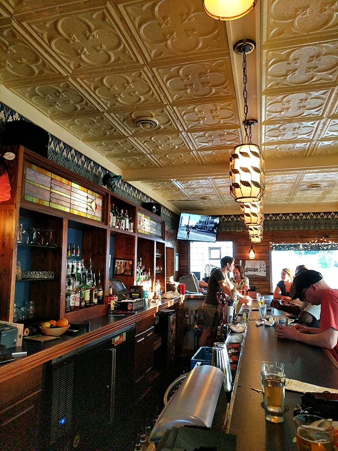 Behind the bar, bottles stand at attention like soldiers guarding liquid treasures, beneath stained glass that filters light into amber jewels.
