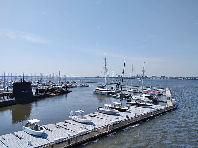 The peaceful marina beside massive warships creates a perfect contrast – sailboats and submarines showing two very different ways to experience water.
