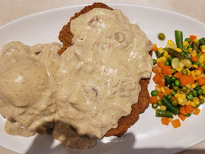 Country fried steak hiding beneath a creamy avalanche of gravy, with vegetables providing a colorful reality check on the side.