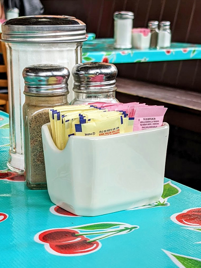 The tabletop essentials: sugar packets, salt, pepper, and a cheerful cherry-patterned tablecloth that whispers, "You're among friends now."