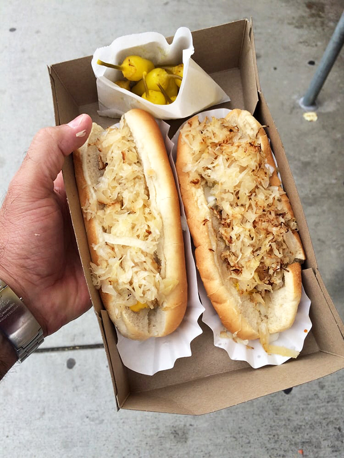 Two dogs nestled in their cardboard cradle, topped with sauerkraut that's been making Californians pretend they're New Yorkers since day one.