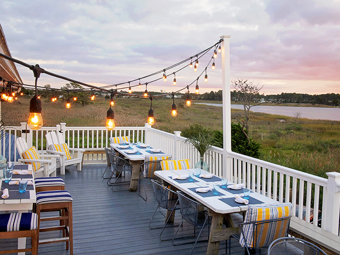 Sunset dining on the deck where string lights twinkle above, marsh grasses sway below, and vacation mode activates with your first sip.