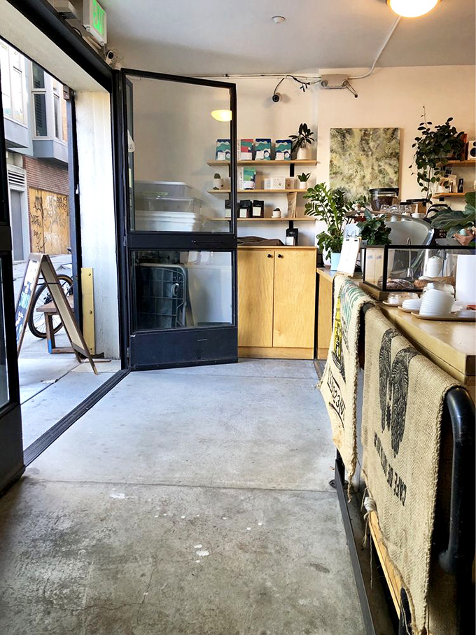Inside, minimalism meets comfort with light wood accents, potted plants, and burlap coffee sacks that create an atmosphere of caffeinated zen.