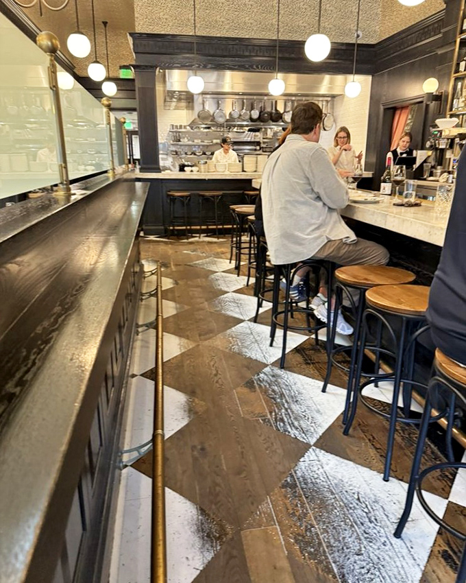 Inside Petit Trois, the classic black and white checkered floor and marble bar create an authentic bistro atmosphere. The open kitchen invites diners to witness culinary artistry in action.