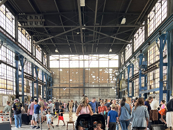 Sunlight streams through massive factory windows, illuminating a bustling marketplace where the ghosts of industry meet modern creativity.