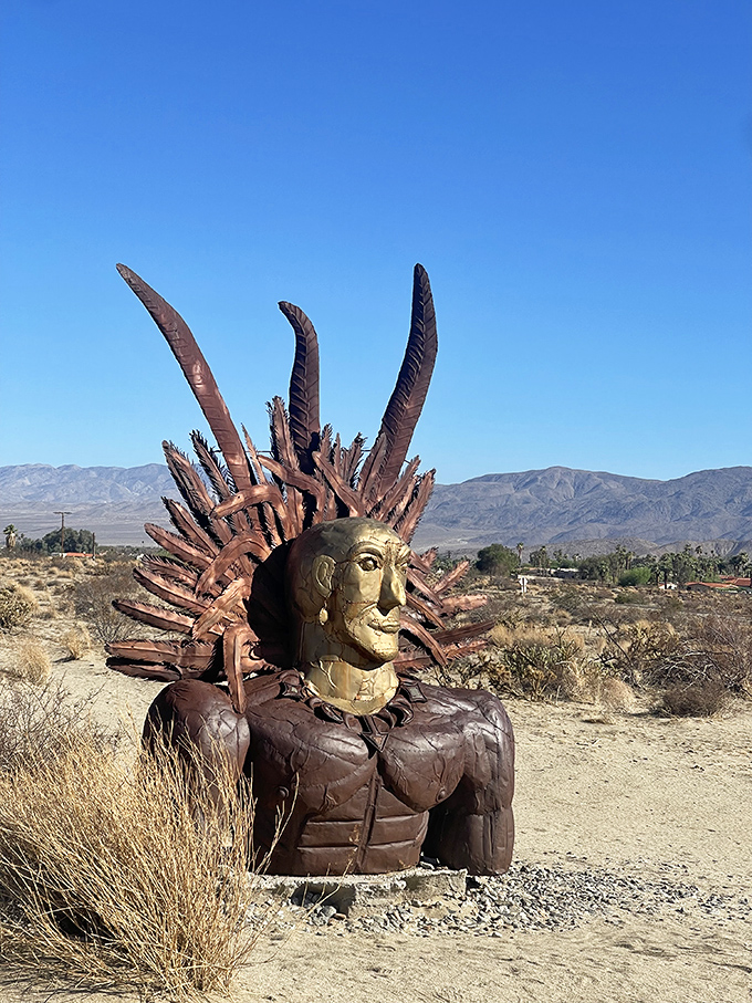 This regal Native American figure stands sentinel in the desert, his golden face catching the sunlight while metal feathers crown his weathered form.