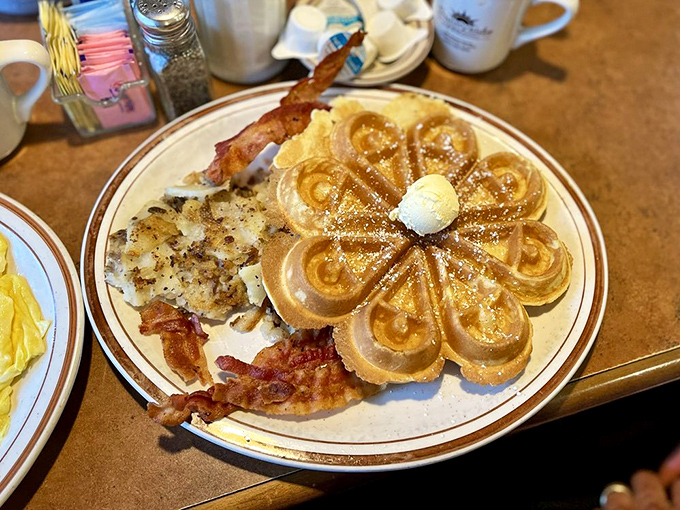 Behold the flower-shaped Belgian waffle in its golden-brown glory, flanked by crispy bacon and hash browns like loyal courtiers to breakfast royalty.
