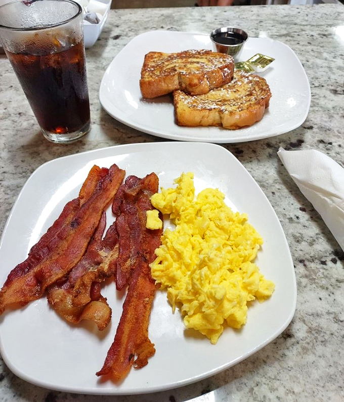 The breakfast trinity: perfectly scrambled eggs that look like sunshine, bacon with the ideal crisp-to-chew ratio, and French toast dusted with powdered sugar.